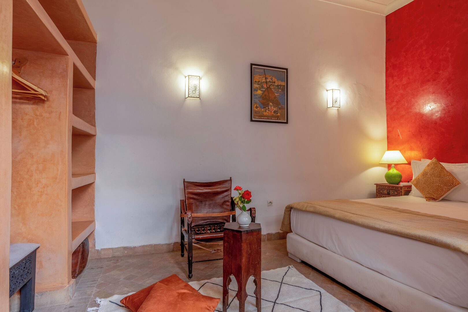 Quarto com cama, uma cadeira, mesa de cabeceira, abajur e um quadro na parede.
