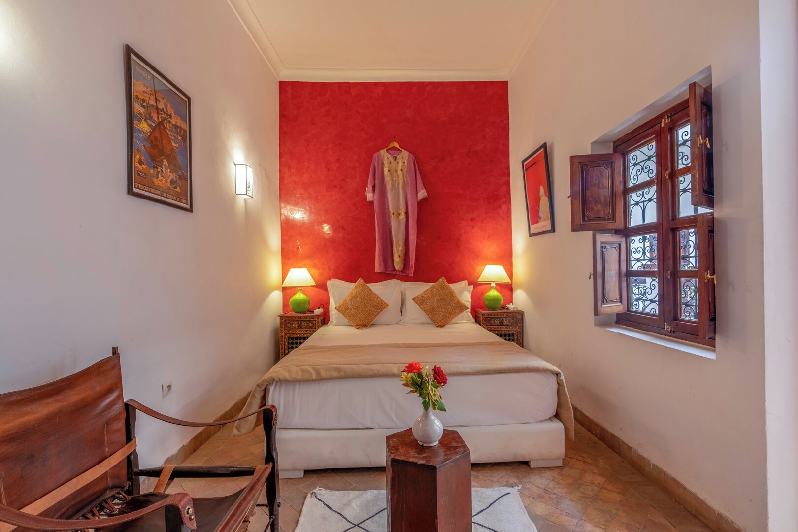 Quarto com parede de destaque vermelha, cama com roupa de cama branca, duas luminárias de cabeceira, uma cadeira de madeira e uma janela com grades de ferro decorativas.