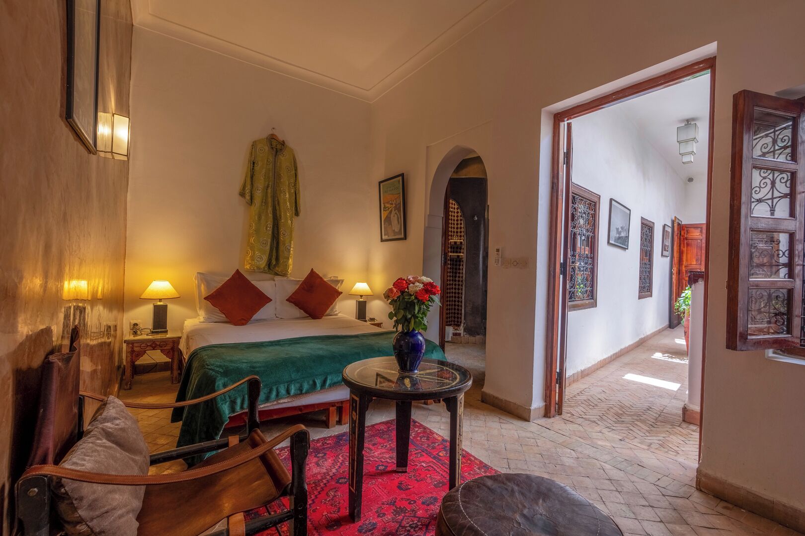 Quarto de hotel com cama, abajures, uma mesa com um vaso, uma cadeira e um tapete vermelho.