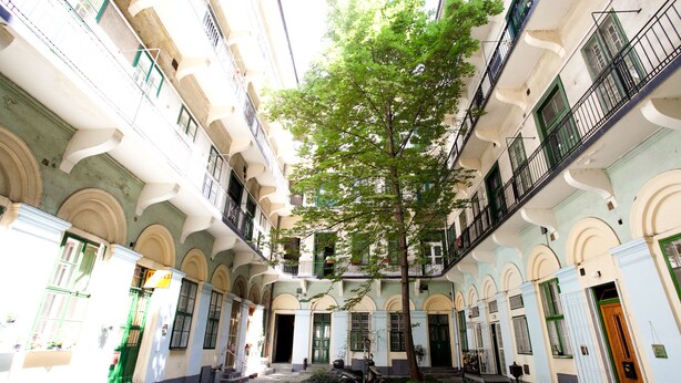 Um pátio com uma árvore, varandas e fachada de edifício com janelas e portas em arco.