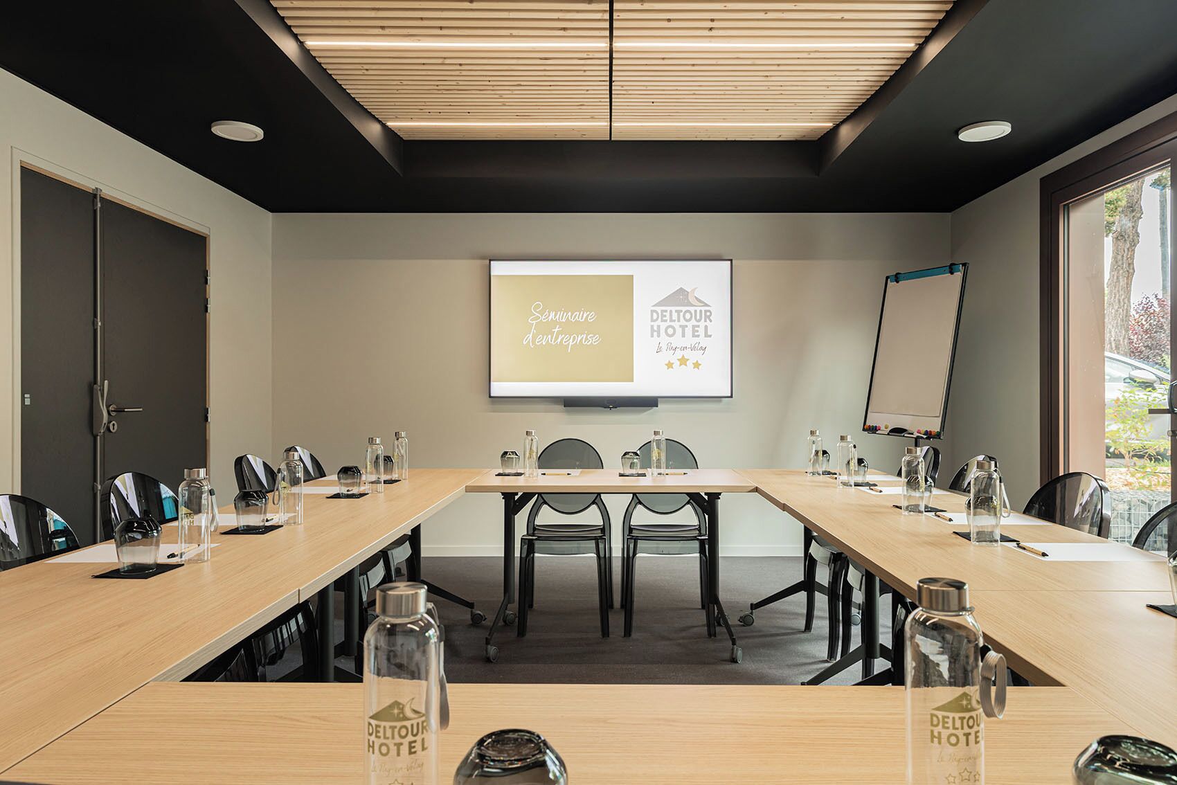 Sala de conferências com uma mesa comprida, cadeiras, tela de projetor e garrafas de água com a etiqueta 'DELTOUR HOTEL'.