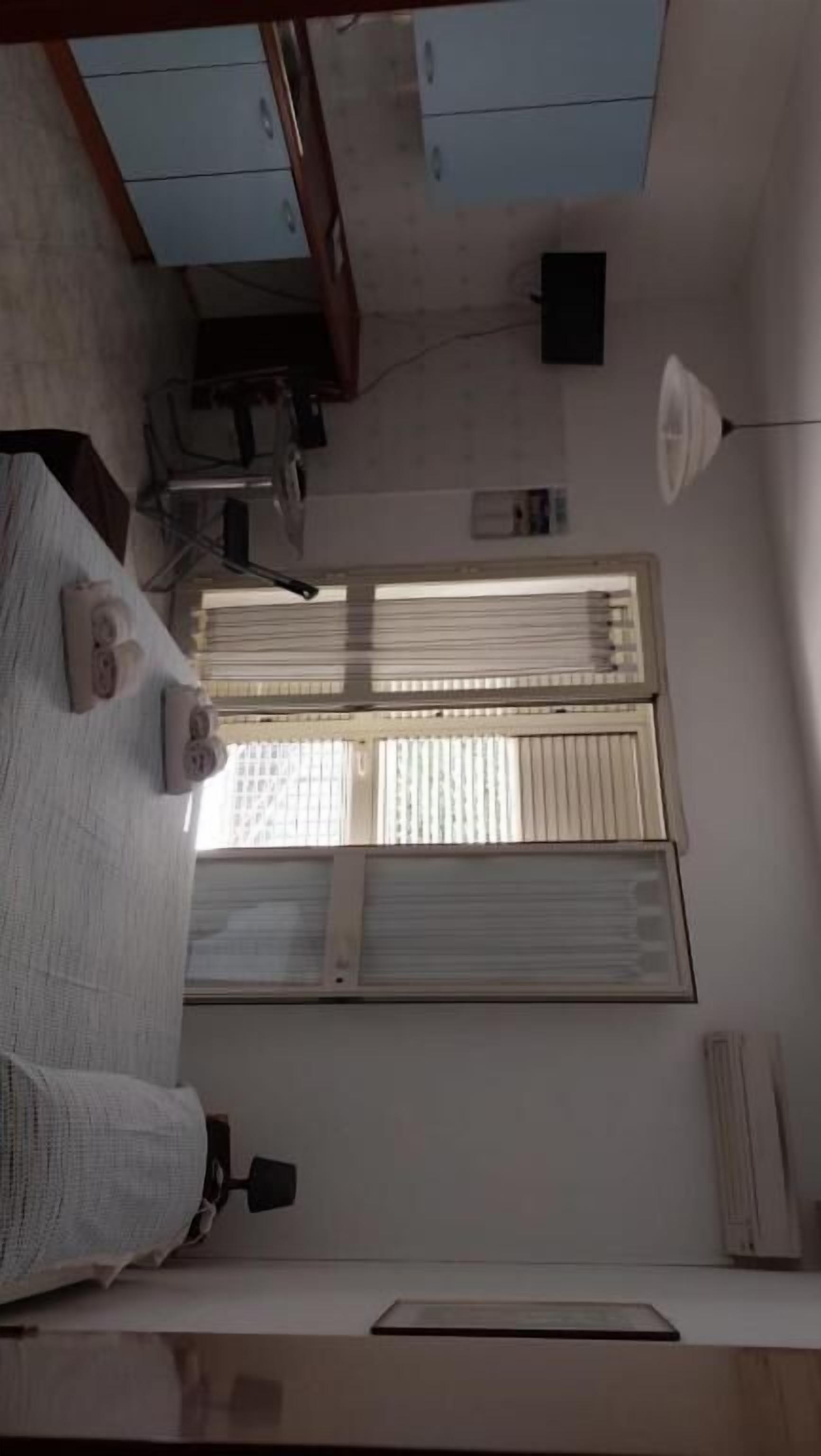 Quarto com cama, escrivaninha, cadeira, janela com persianas, radiador e abajur.