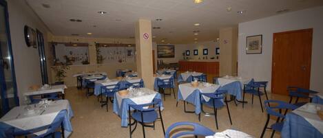 Uma sala de jantar com mesas preparadas para o serviço, cadeiras azuis e um buffet ao fundo.