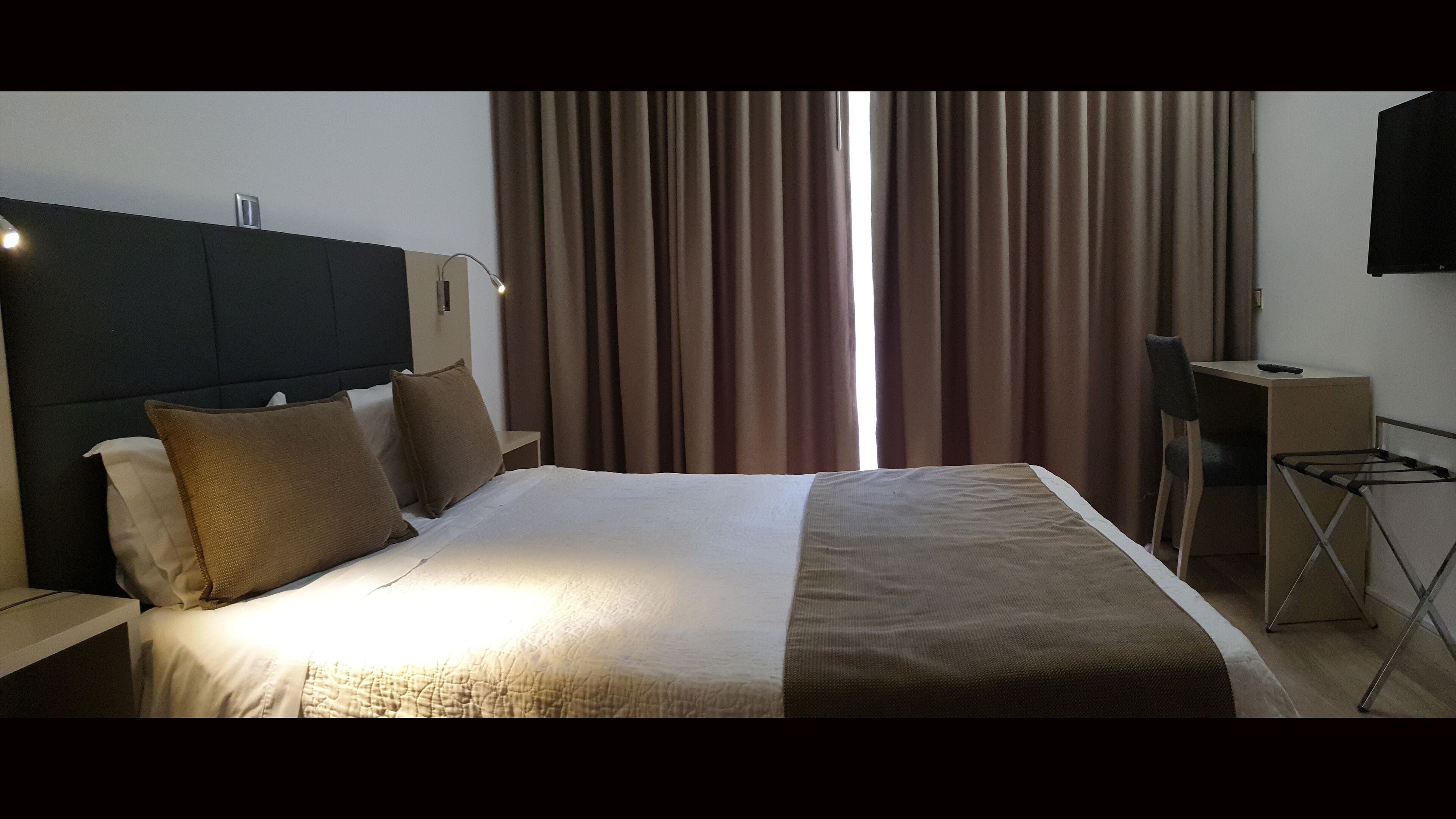 Quarto de hotel com cama, escrivaninha, cadeira e televisão.