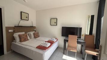 Quarto de hotel com uma cama, escrivaninha com computador, cadeira e duas televisões na parede.