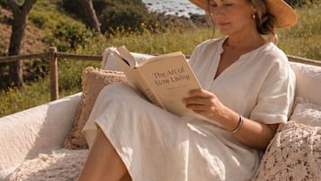 Uma pessoa sentada em um sofá com um livro intitulado 'The Art of Slow Living' em um ambiente natural, com árvores e vista para o mar.