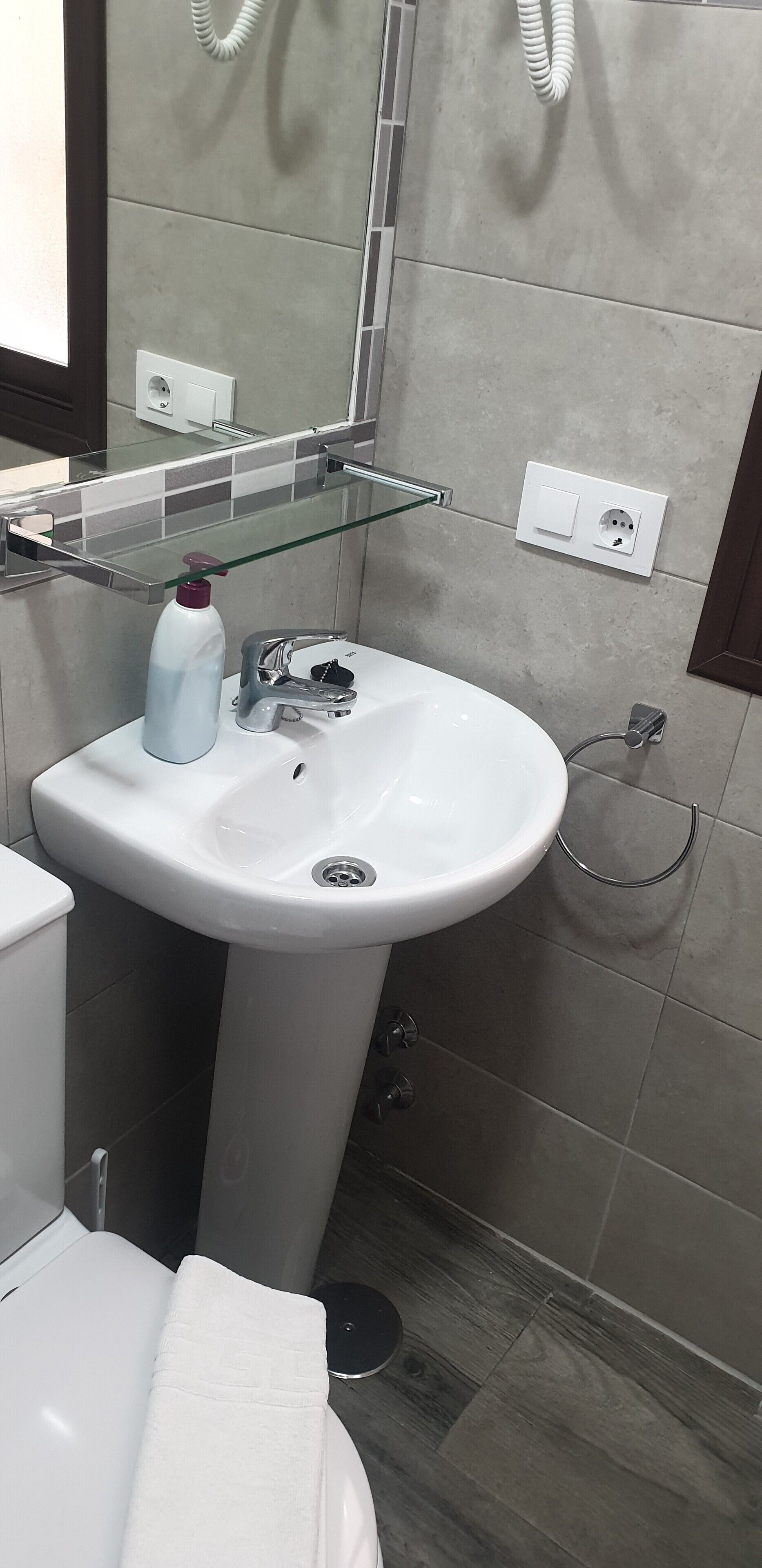 Casa de banho com uma pia branca, uma prateleira de vidro, uma garrafa de sabonete líquido e uma toalha no sanitário.