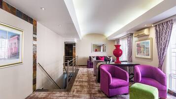 Quarto de hotel com escada, área de estar com poltronas roxas, um banquinho verde e um vaso vermelho sobre uma mesa.