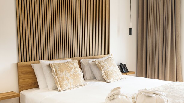 Quarto de hotel com cabeceira de madeira, cama com roupa de cama branca e bege, duas almofadas, mesa de cabeceira com telefone e cortina.