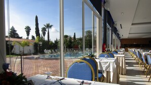 Uma área de refeições com mesas preparadas para hóspedes, grandes janelas com vista para a piscina e palmeiras, e um céu claro.