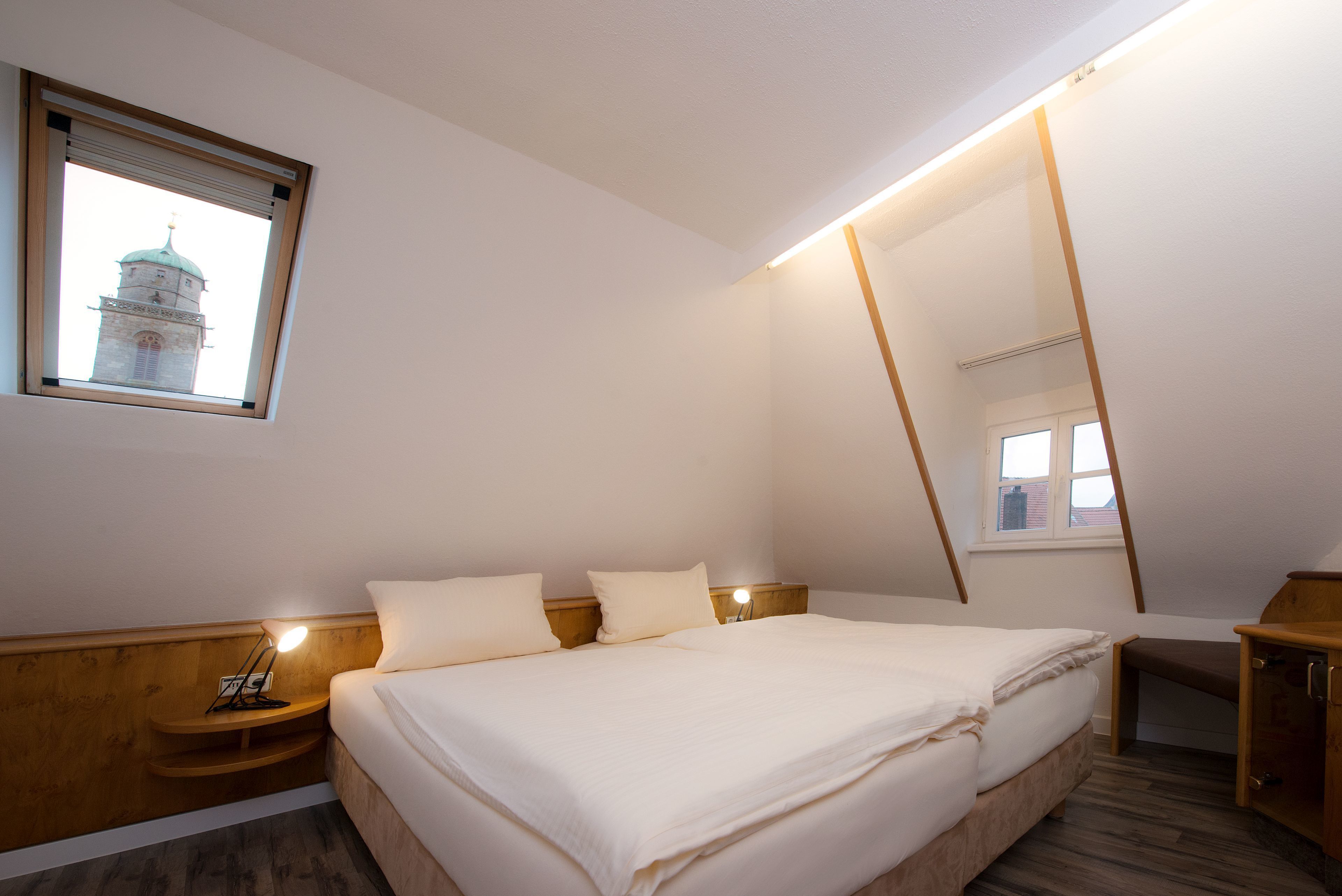 Quarto com cama, mesas de cabeceira, um criado-mudo, janela com vista para uma torre e teto inclinado.