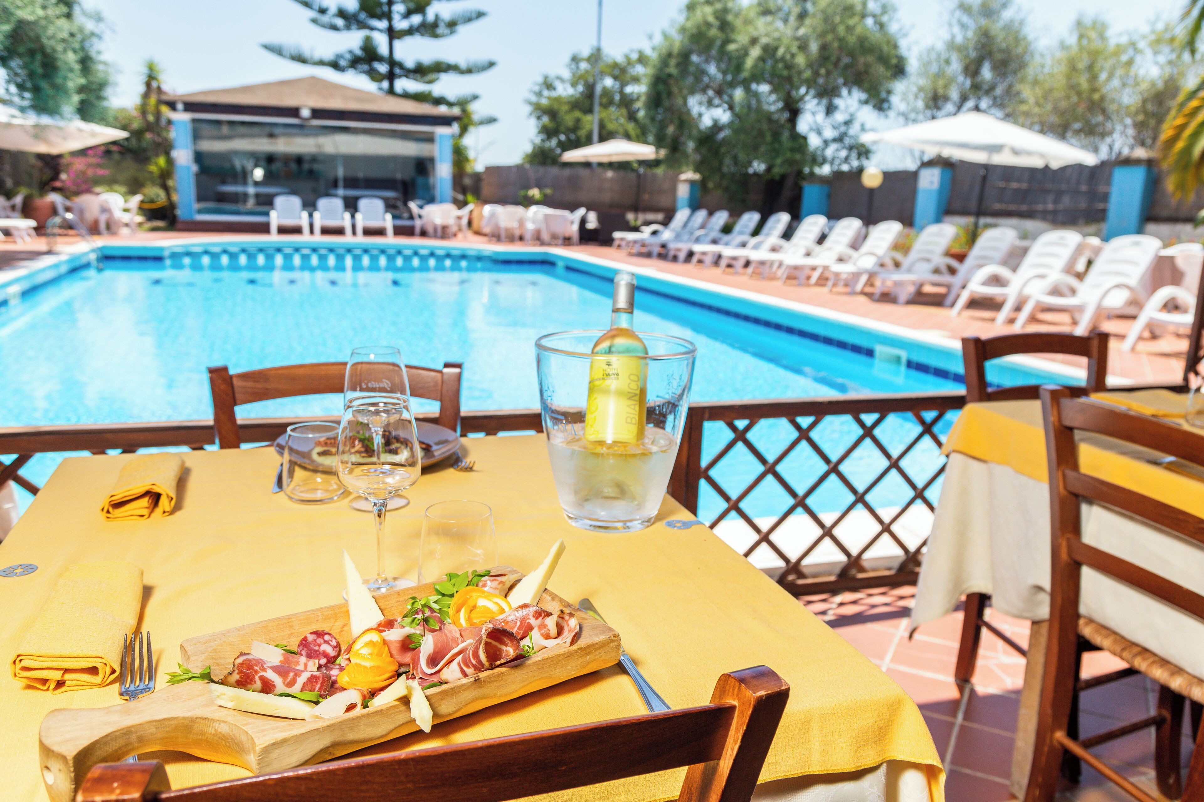 Mesa de jantar com refeição, uma garrafa de vinho e um copo de água, com vista para uma piscina e espreguiçadeiras.
