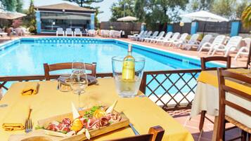Mesa de jantar com refeição, uma garrafa de vinho e um copo de água, com vista para uma piscina e espreguiçadeiras.