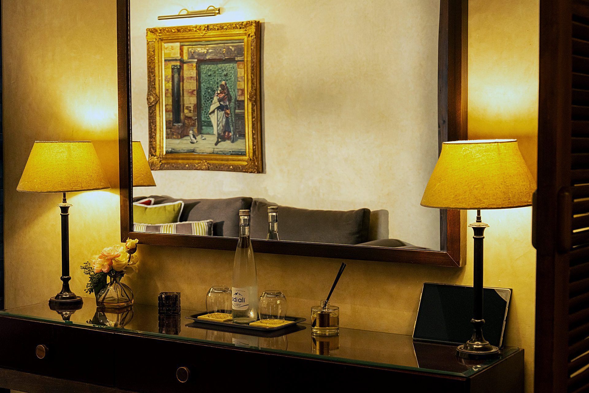 Quarto de hotel com espelho, duas luminárias, um vaso com flores, um tabuleiro com copos e um quadro na parede.