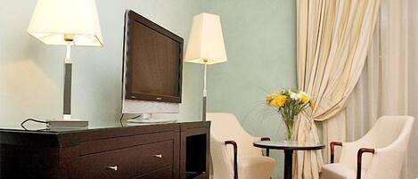 Quarto de hotel com cômoda de madeira escura, duas luminárias, TV de tela plana, mesa redonda pequena com um vaso de flores, duas cadeiras brancas e uma janela grande com cortinas bege.