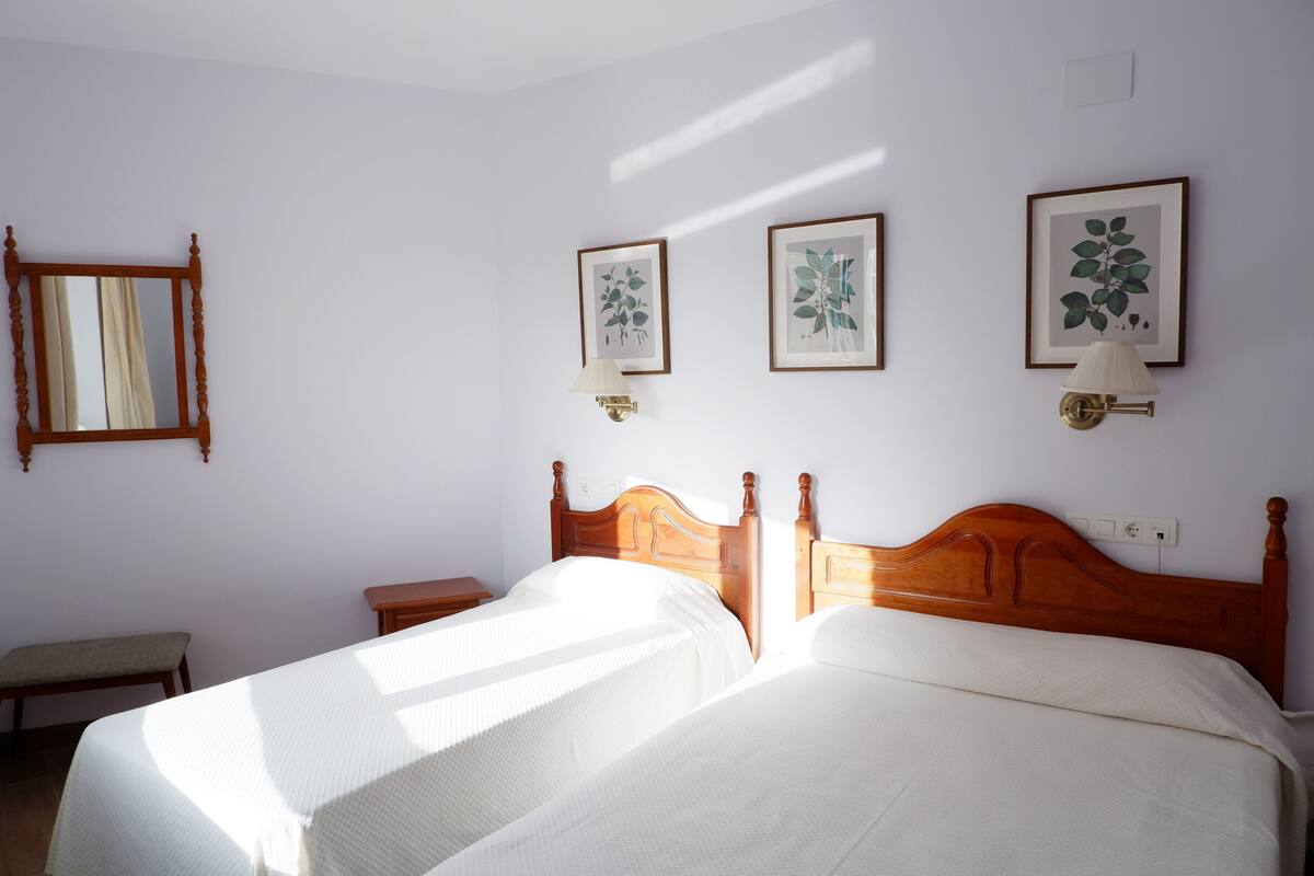 Quarto de hotel com duas camas, cabeceira de madeira, mesa de cabeceira, um banquinho e obras de arte na parede.
