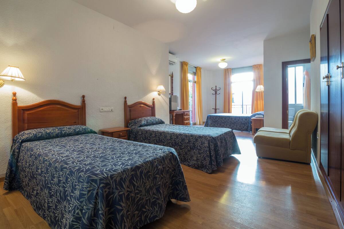 Quarto de hotel com duas camas, piso de madeira, uma cadeira e uma escrivaninha com abajur.