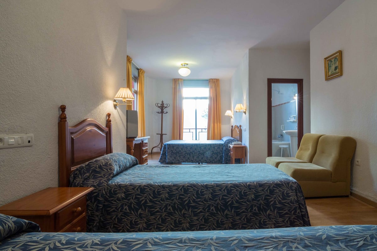 Quarto de hotel com duas camas, uma escrivaninha, uma cadeira e uma mesinha.