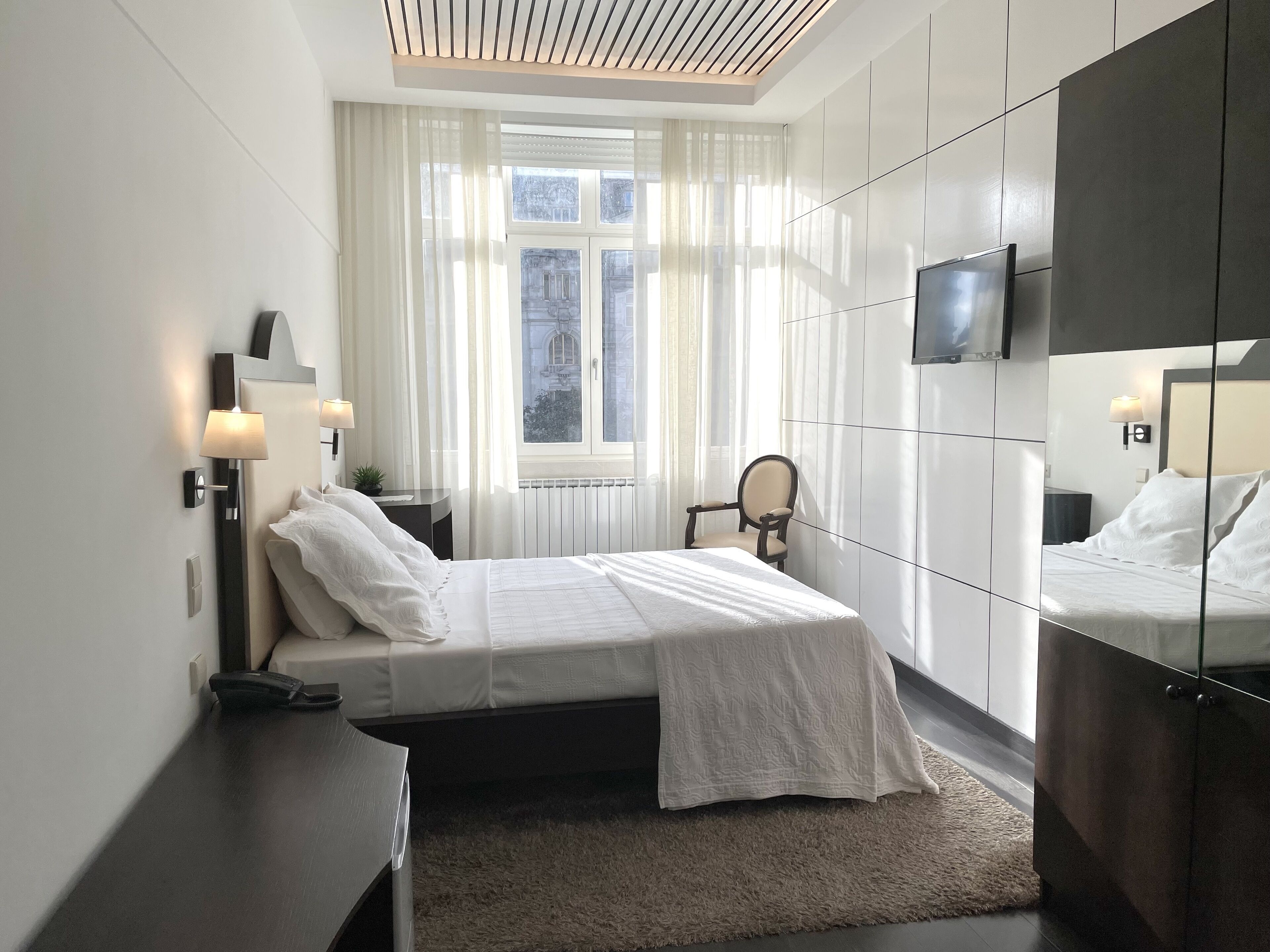 Quarto de hotel moderno com uma cama grande, televisão de tela plana, espelho e janela com cortinas leves.