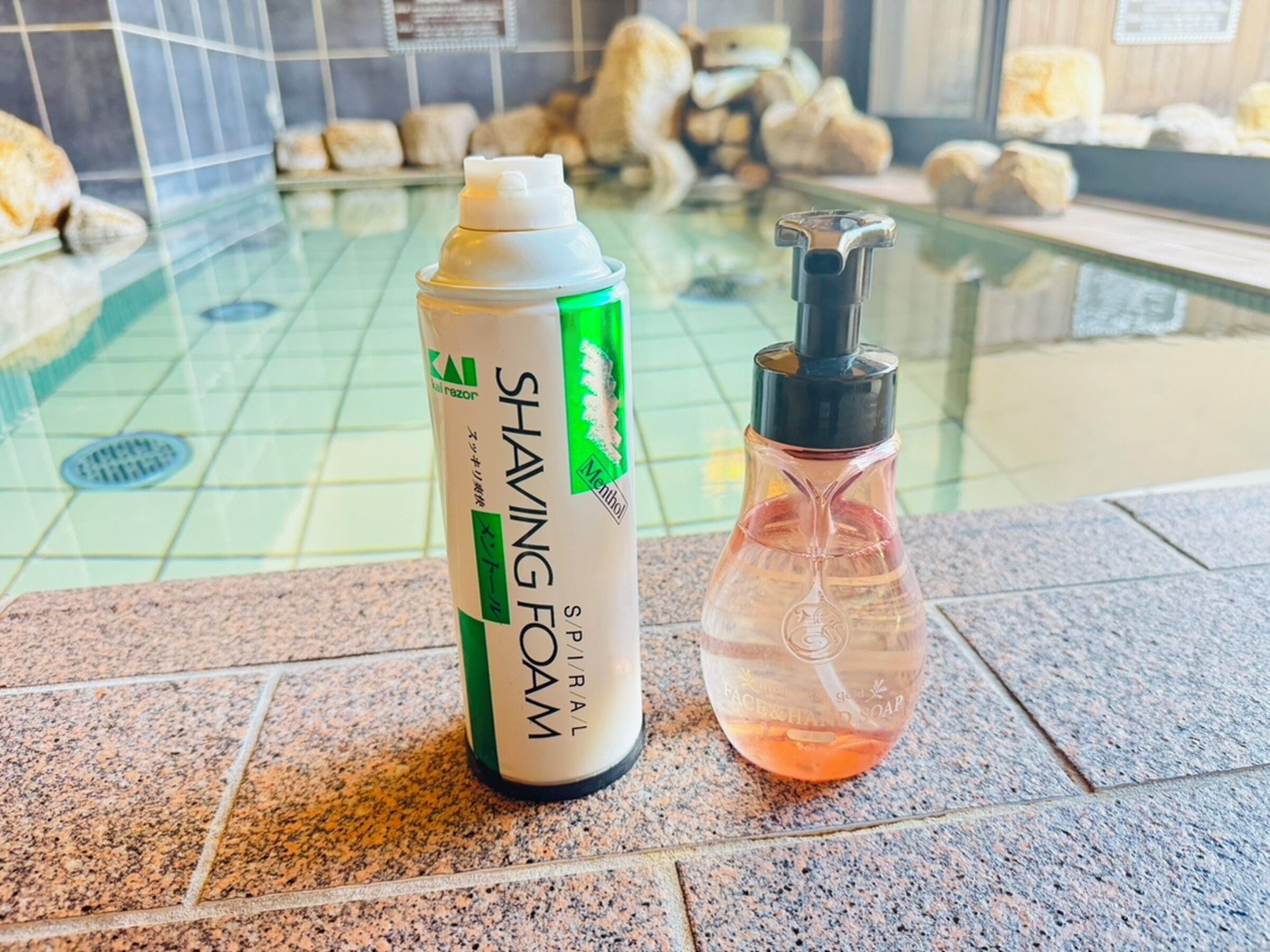Uma embalagem de espuma de barbear e um frasco dosador de sabonete líquido sobre uma superfície azulejada perto de uma piscina.