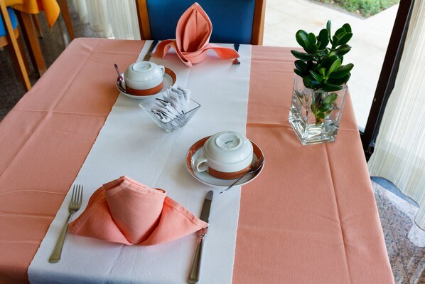 Uma mesa de jantar bem posta, com toalhas de mesa brancas e cor-de-pêssego, dois bules brancos com pires, uma tigela de vidro com embalagens, um guardanapo rosa, um garfo e uma faca.