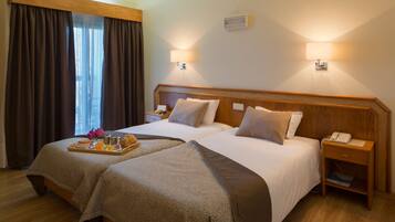 Quarto de hotel com duas camas, cabeceira de madeira, mesas de cabeceira com abajures, bandeja com itens de café da manhã e uma janela com cortinas.