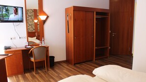Quarto de hotel com escrivaninha de madeira, cadeira, cama, guarda-roupa e televisão presa na parede.