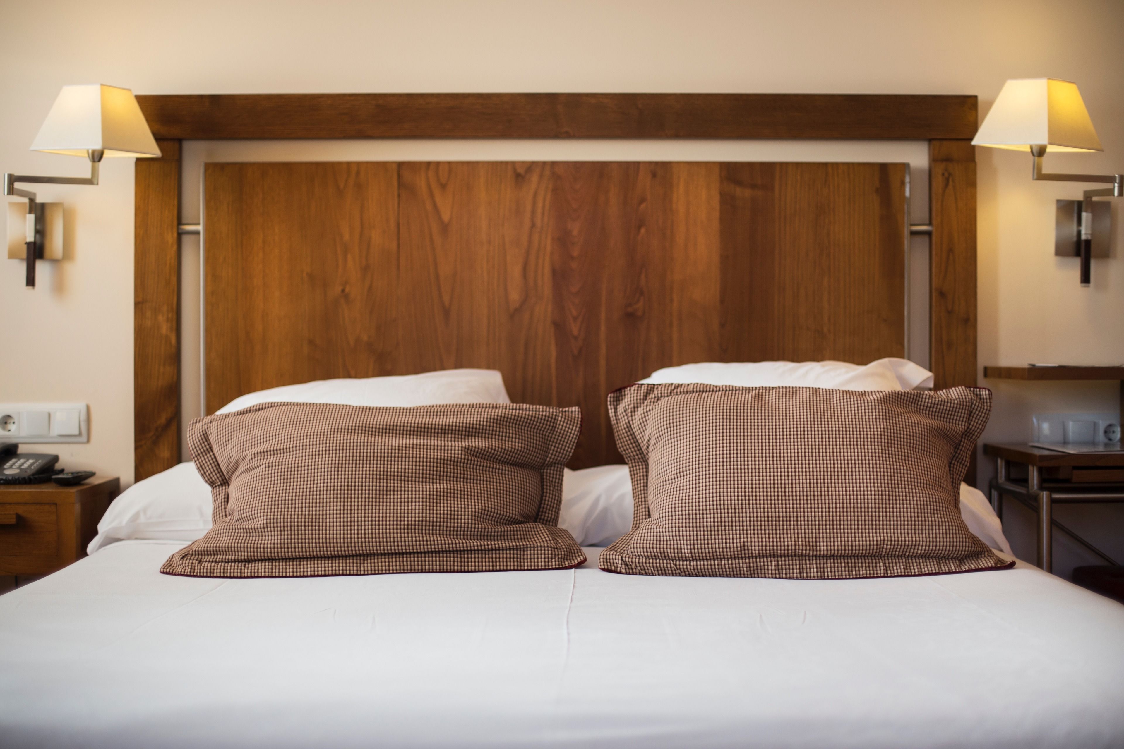 Quarto de hotel com cabeceira de madeira, duas almofadas xadrez, roupa de cama branca e luminárias fixadas na parede.