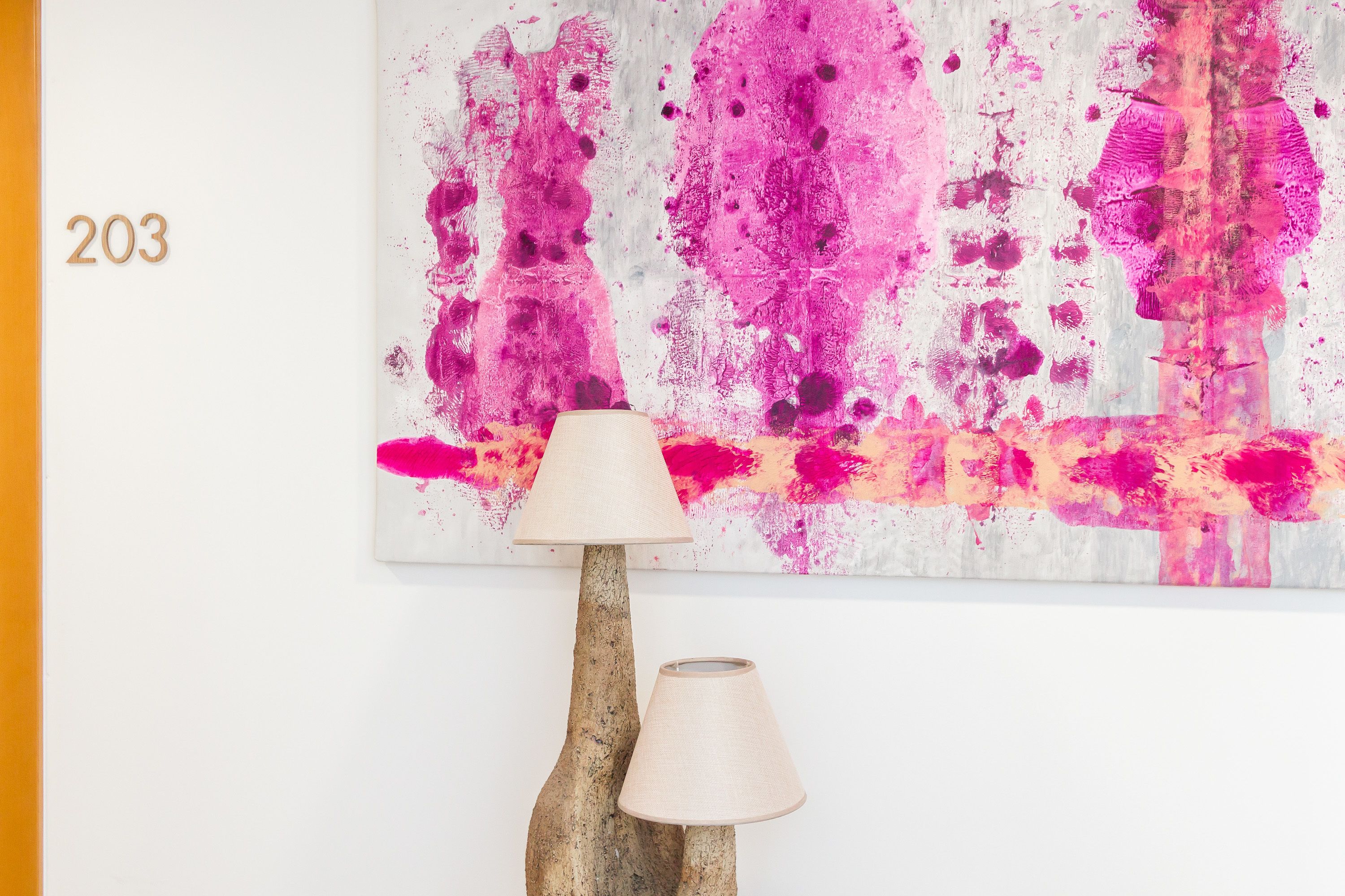 Quarto com o número '203' na parede, uma pintura com salpicos abstratos em rosa e branco, e duas luminárias com cúpulas bege.