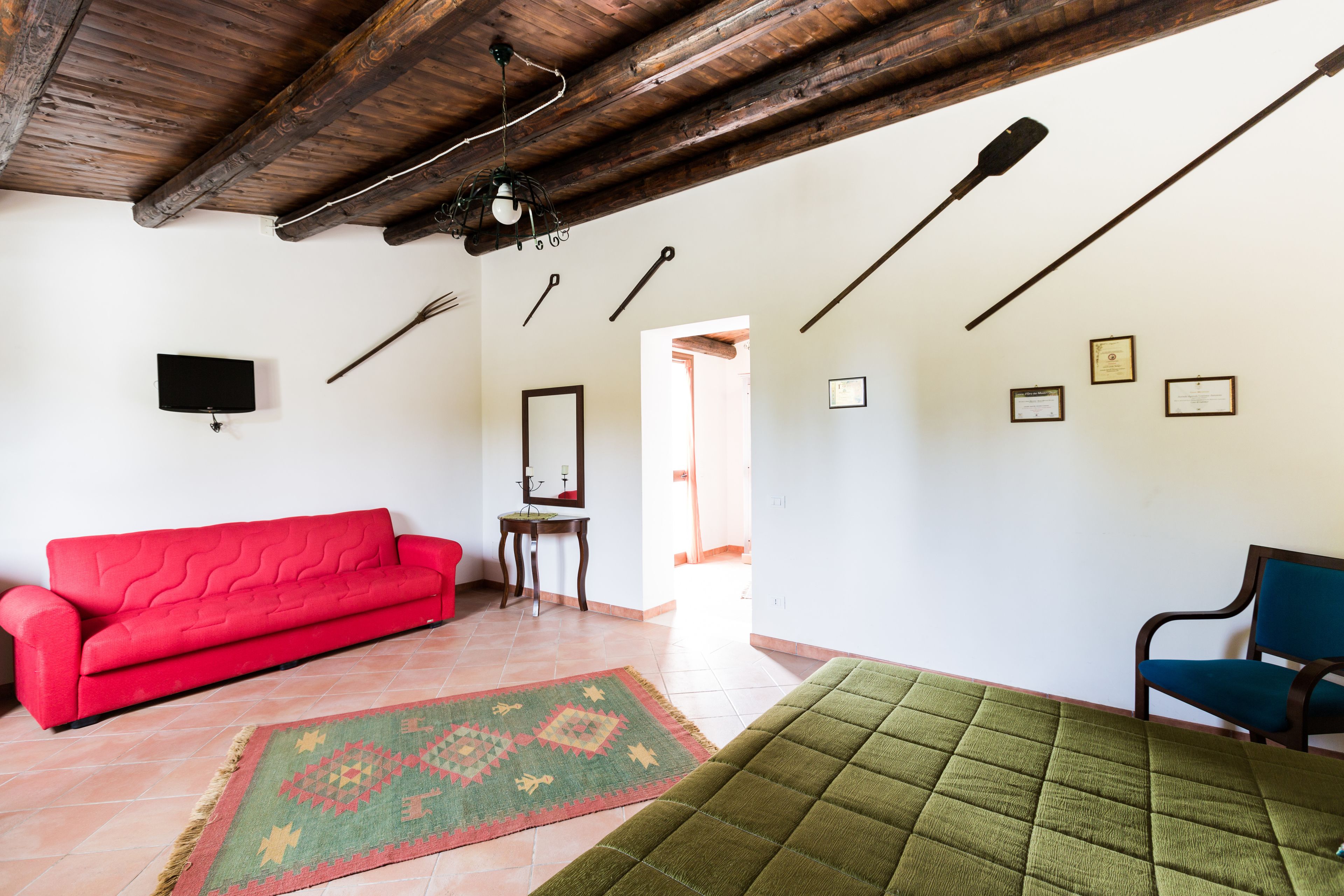 Quarto com sofá vermelho, um banquinho verde, uma cadeira azul e teto de madeira com vigas aparentes.