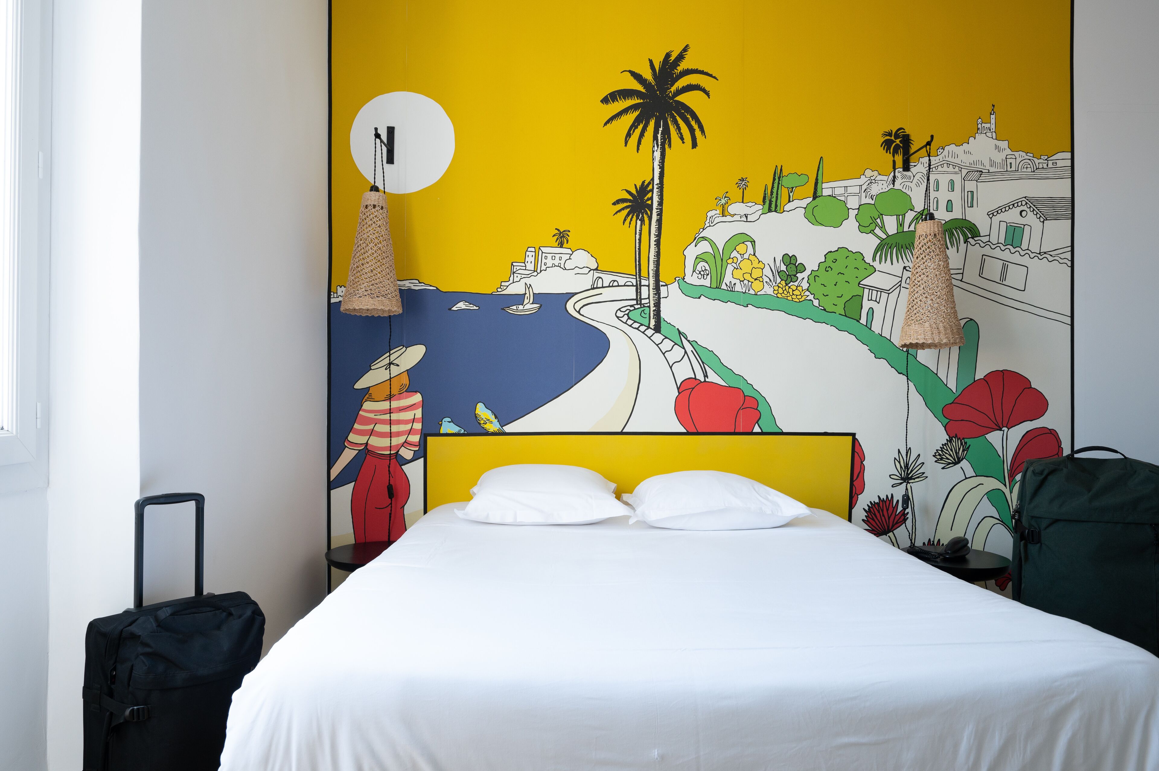 Quarto de hotel com cama, mala e um mural vibrante na parede retratando uma paisagem costeira.