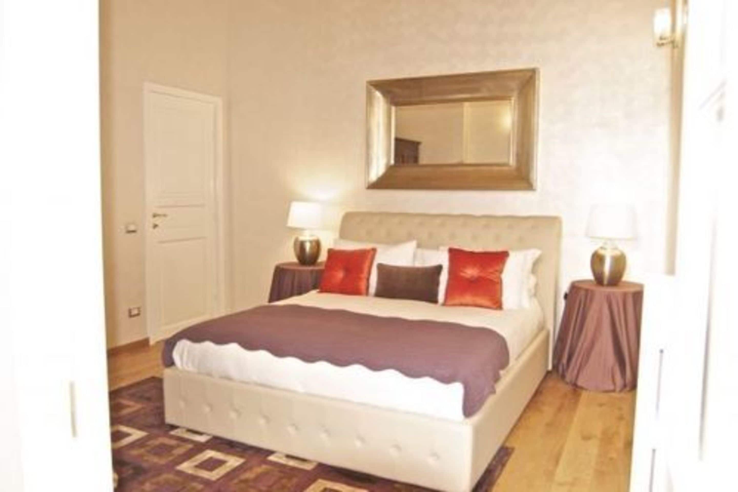 Quarto com uma cama grande, duas mesas de cabeceira com abajures, um espelho sobre a cama e uma porta à esquerda.