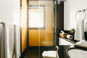 Uma casa de banho com cabine de duche em vidro, sanita, lavatório com toalha e suporte para papel higiénico.