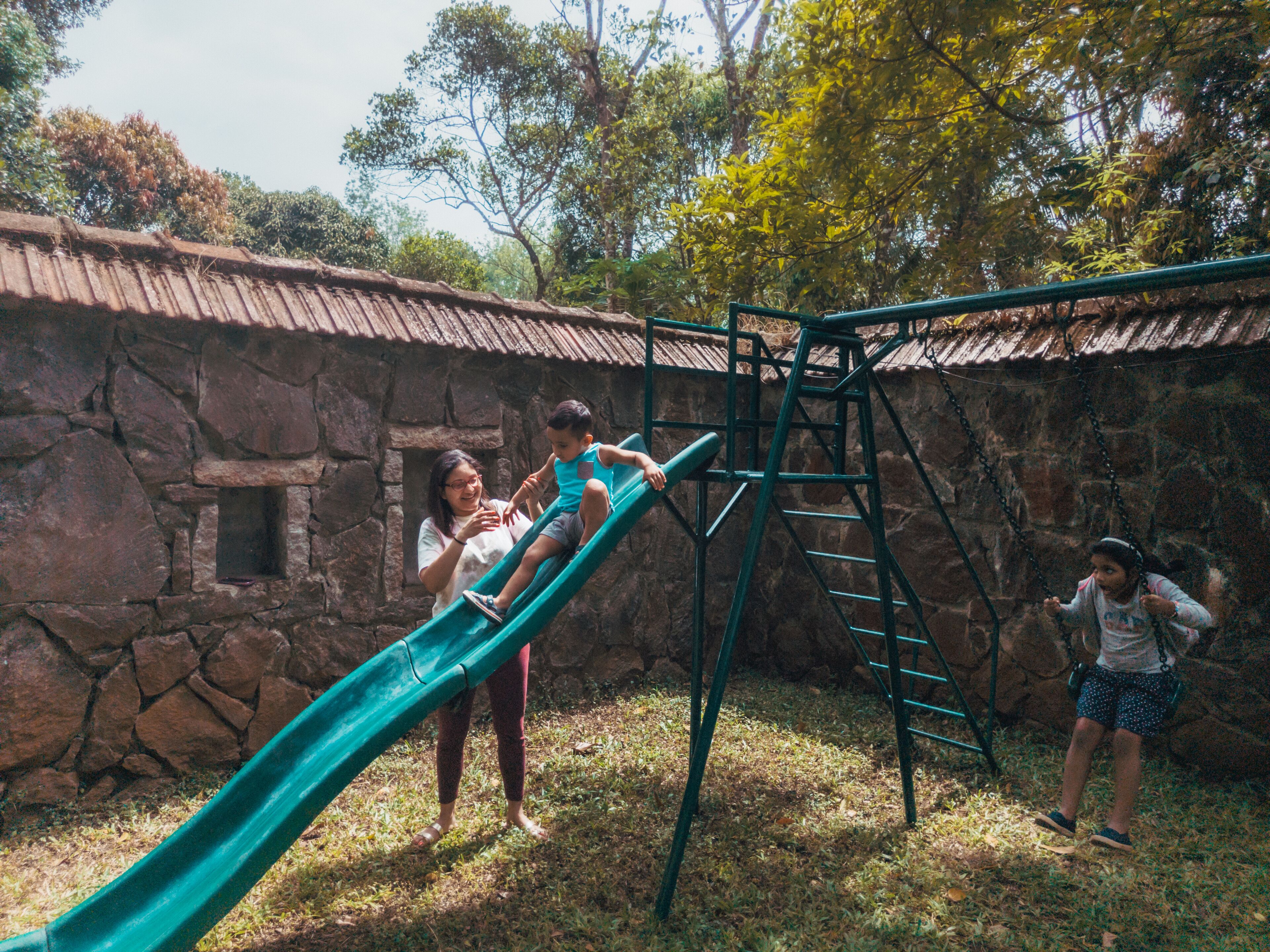 Um parquinho com um escorrega verde, uma parede de pedra e uma escada de metal.