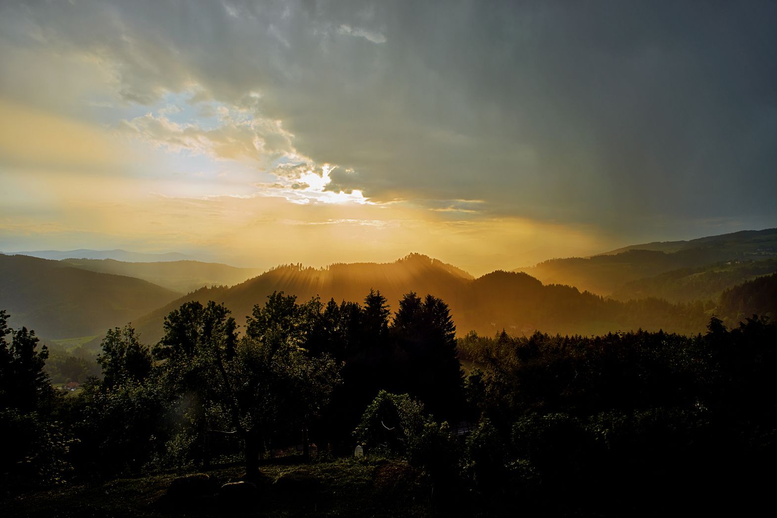 Paisagem montanhosa com nascer ou pôr do sol, destacando colinas onduladas e uma floresta densa em primeiro plano.