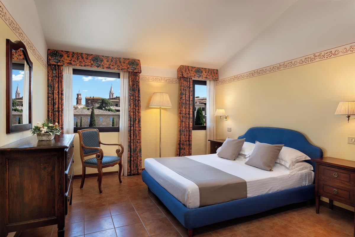 Quarto de hotel com cama, escrivaninha, cadeira, luminária e vista para edifícios pela janela.