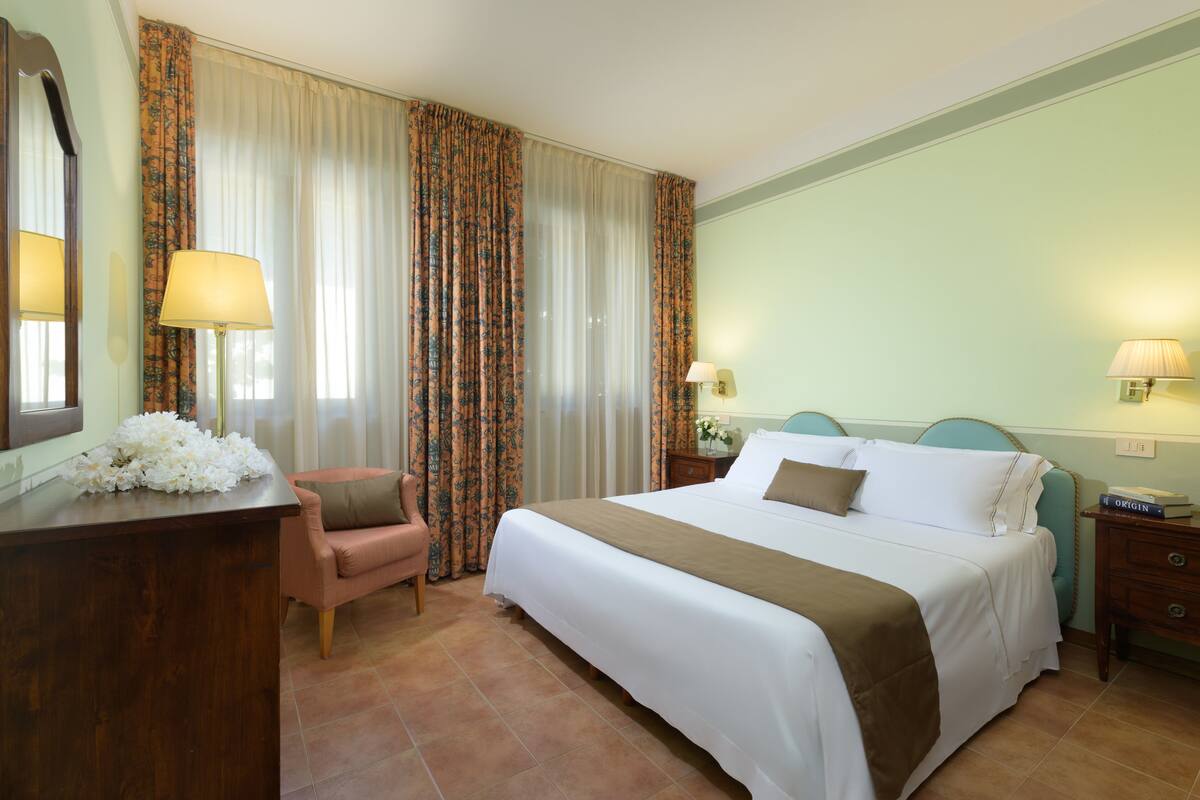 Quarto de hotel com uma cama, uma escrivaninha, uma cadeira e cortinas.