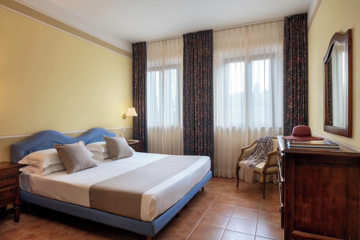 Quarto de hotel com uma cama, escrivaninha, cadeira, abajur e espelho.