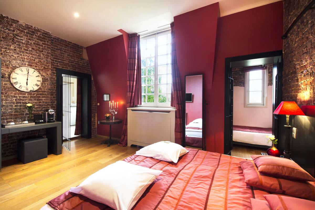 Quarto com parede de tijolos, uma janela grande com cortinas, cama com roupa de cama vermelha, mesa de cabeceira com abajur e um relógio na parede.