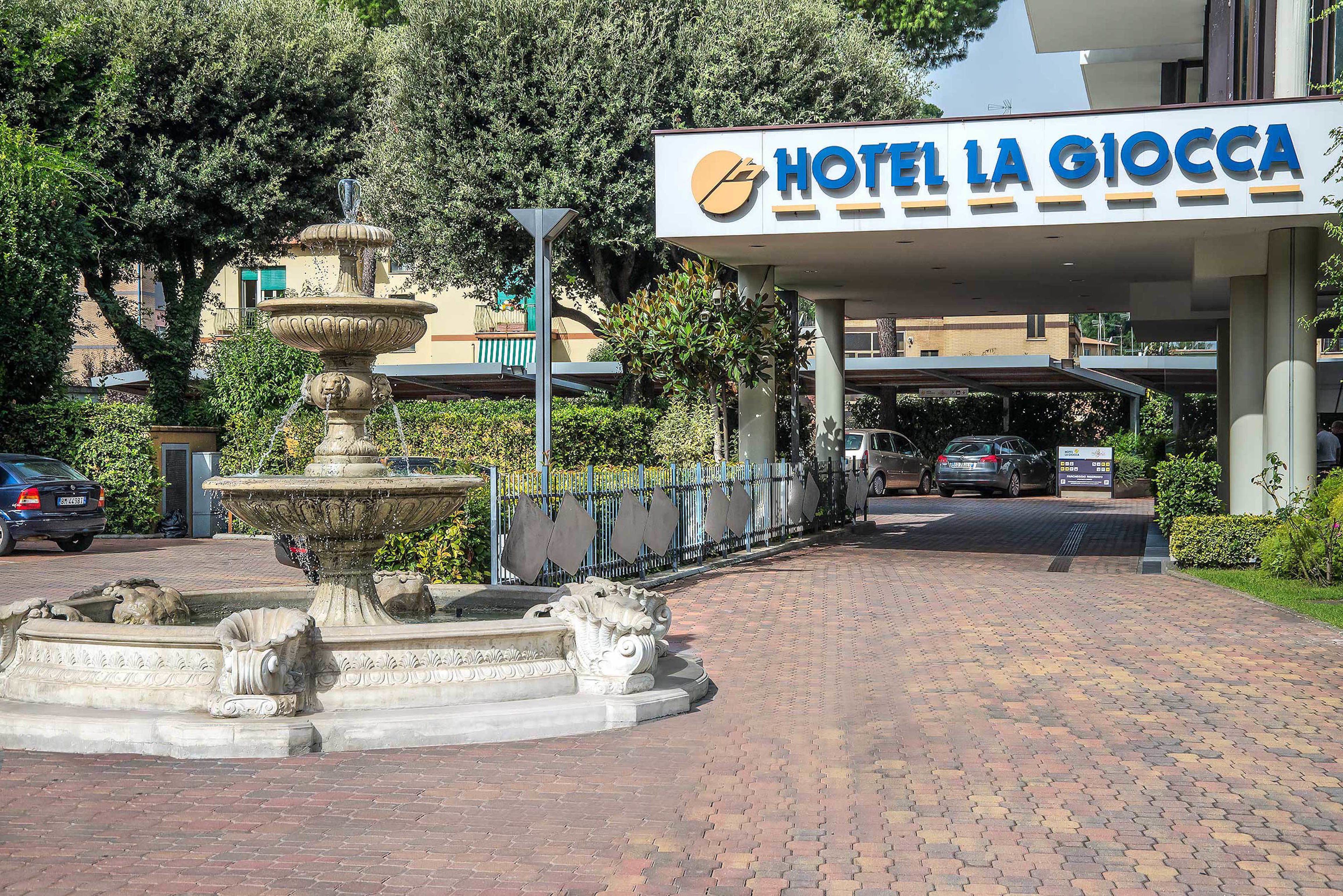 Entrada de hotel com uma fonte, uma placa com a inscrição "HOTEL LA GIOCCA" e um acesso pavimentado.