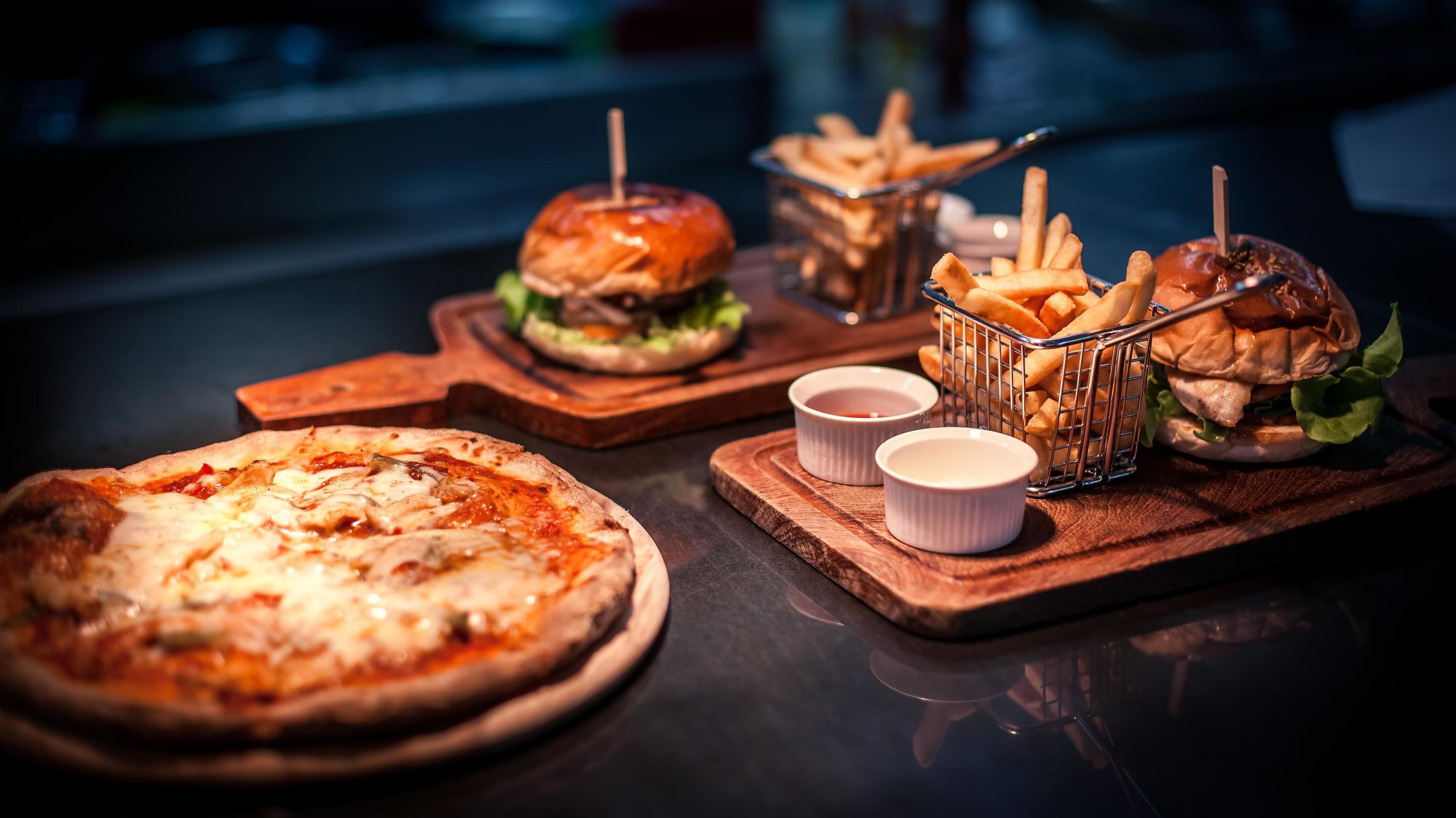 Uma mesa de madeira com pizza, hambúrguer, batatas fritas e uma bebida.