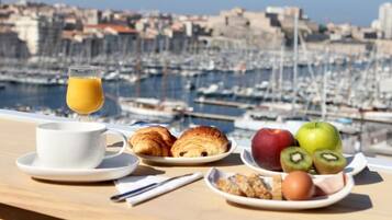 Mesa de café da manhã com croissants, frutas e um copo de suco de laranja em uma varanda com vista para um porto.