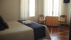 Quarto de hotel com cama, duas cadeiras, uma mesa pequena e uma televisão.