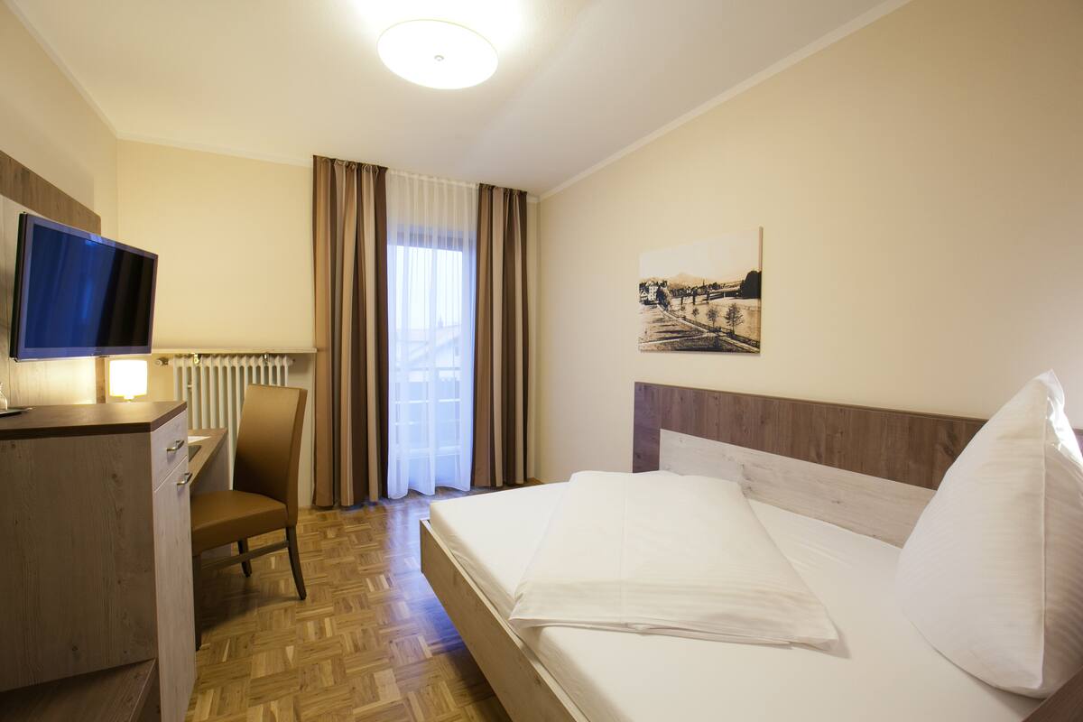 Quarto de hotel com cama, escrivaninha com televisão, cadeira e janela com cortinas.