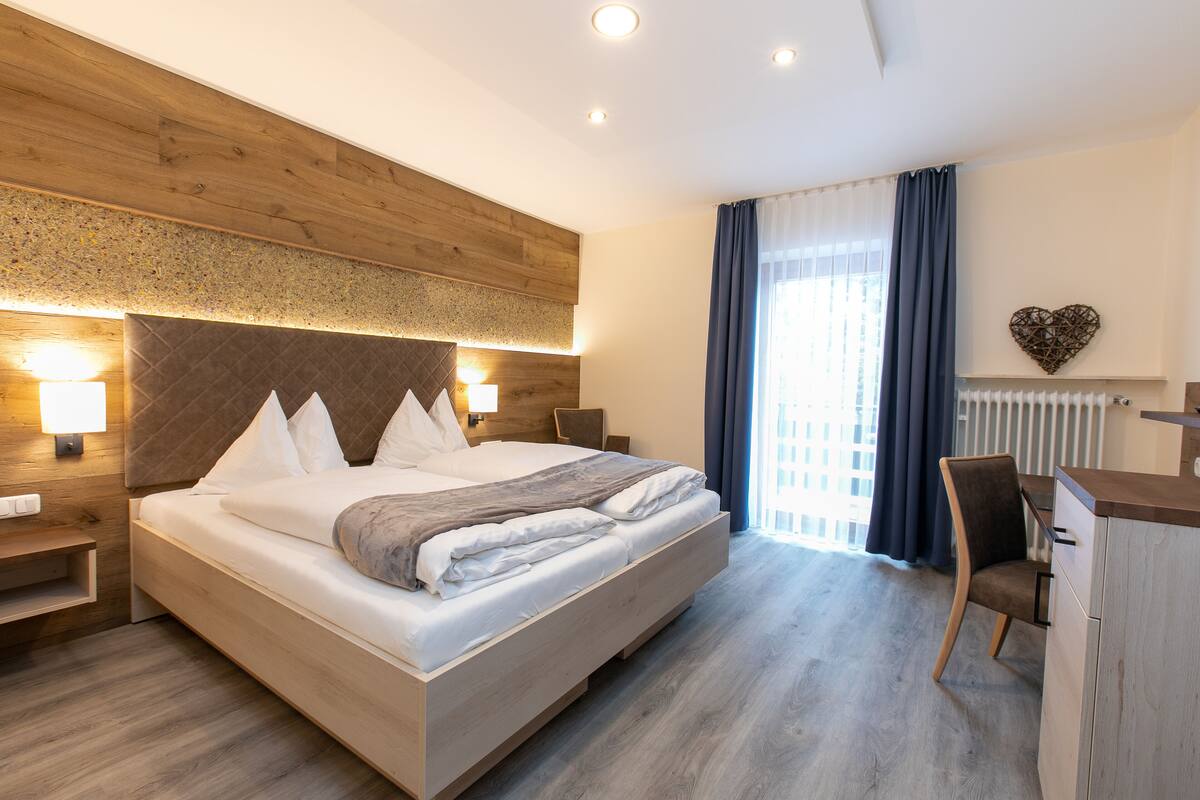 Quarto de hotel moderno com uma cama grande, cabeceira de madeira, abajures, uma escrivaninha e uma cadeira.