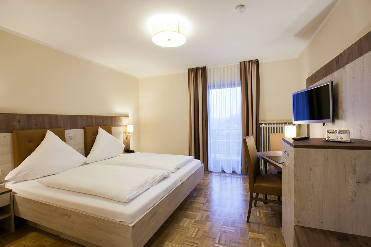 Quarto de hotel com cama de casal, escrivaninha com computador, televisão e janela com cortinas.