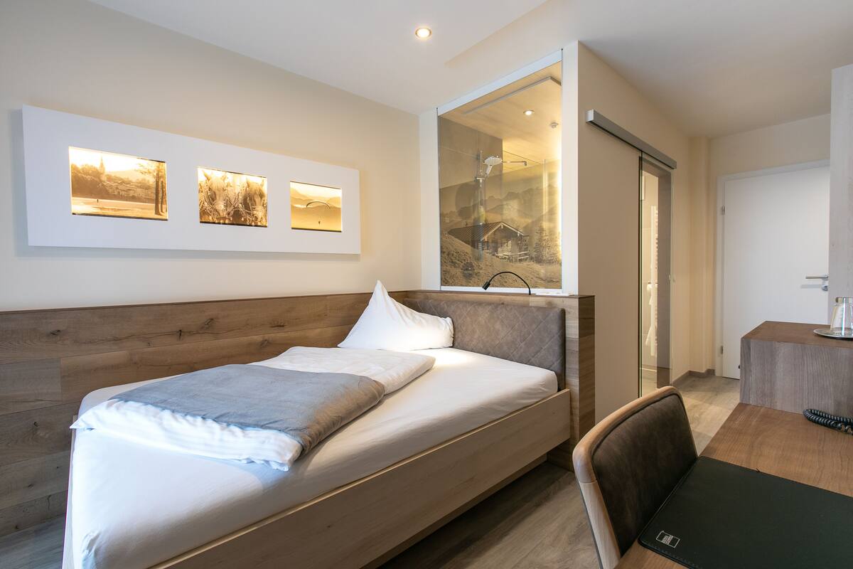 Quarto de hotel moderno com cama de madeira, escrivaninha e um espelho grande refletindo o aposento.
