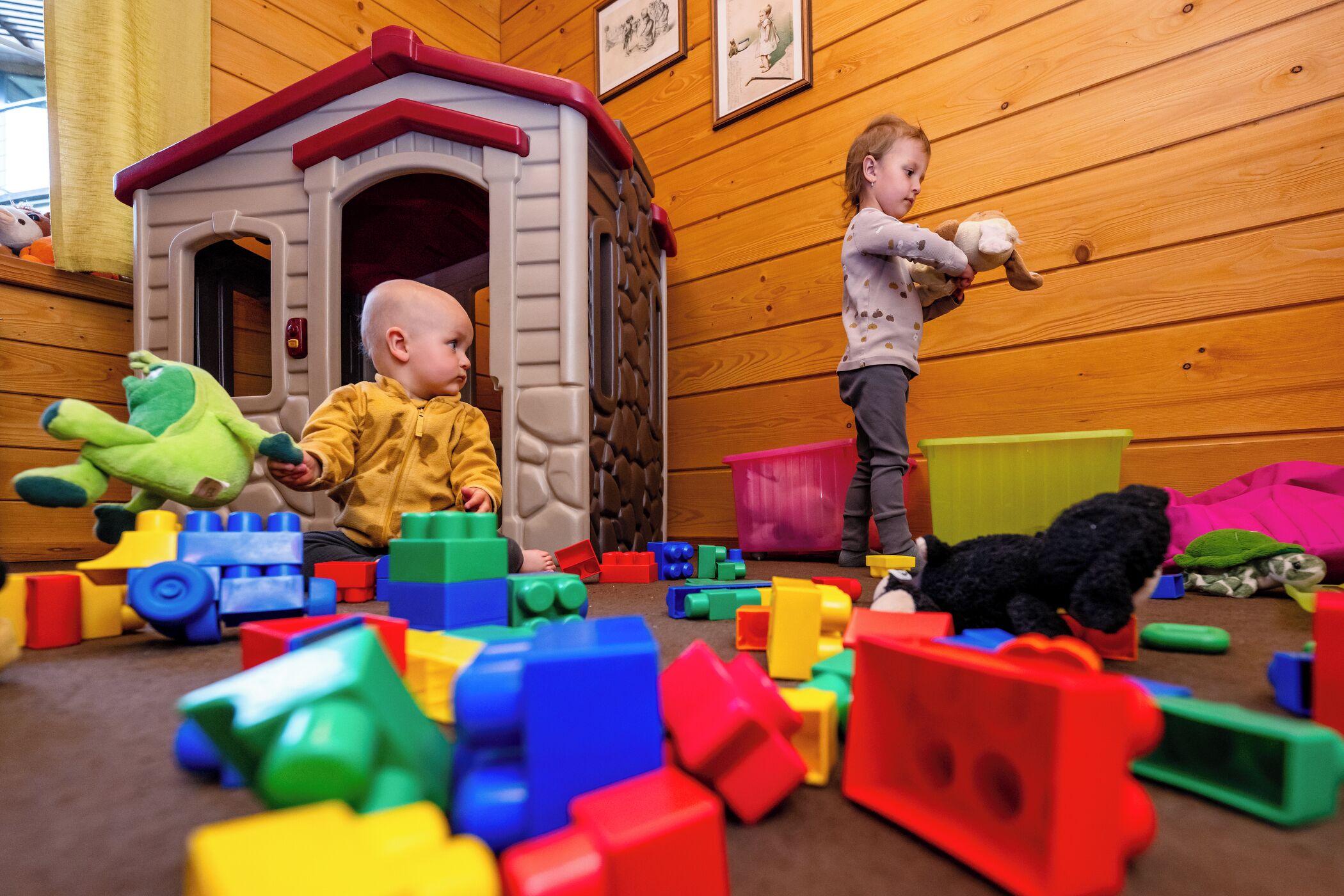 Crianças brincando com blocos de construção coloridos em um quarto com paredes de madeira e uma casinha.