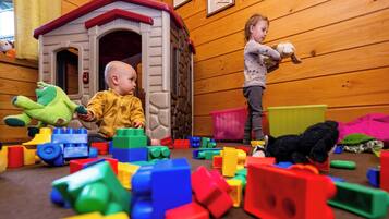 Crianças brincando com blocos de construção coloridos em um quarto com paredes de madeira e uma casinha.