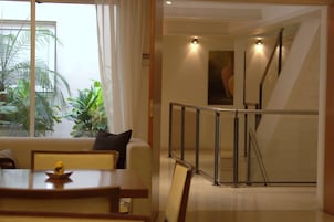 Um saguão de hotel moderno com uma escadaria, uma área de refeições com mesa e cadeiras, e uma janela grande com plantas.